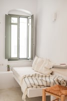 Quarto minimalista com cama, janela com persianas verdes, uma planta pequena e uma mesa de cabeceira com um livro.