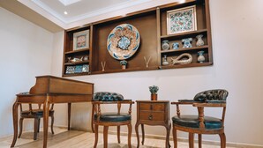 Quarto com piano, duas cadeiras, uma mesinha e uma estante decorativa com vasos e uma obra de arte central.