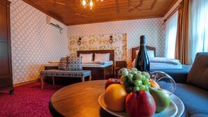 Quarto de hotel com teto de madeira, cama com cabeceira, mesa de cabeceira com uma garrafa, uma cadeira, mesa redonda com frutas e um tapete estampado.