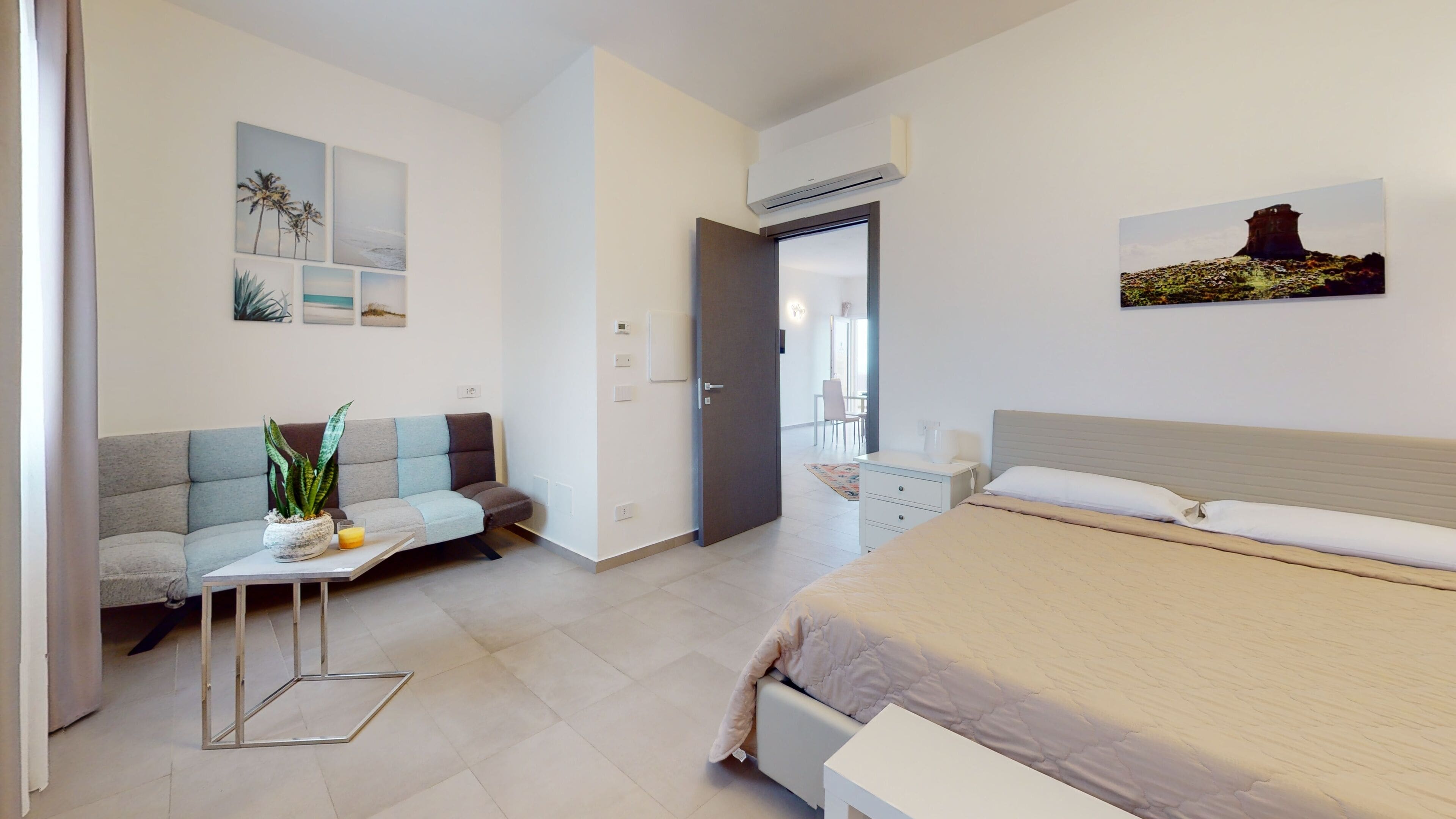 Quarto de hotel moderno com uma cama, um sofá, uma mesinha com uma planta e obras de arte nas paredes.
