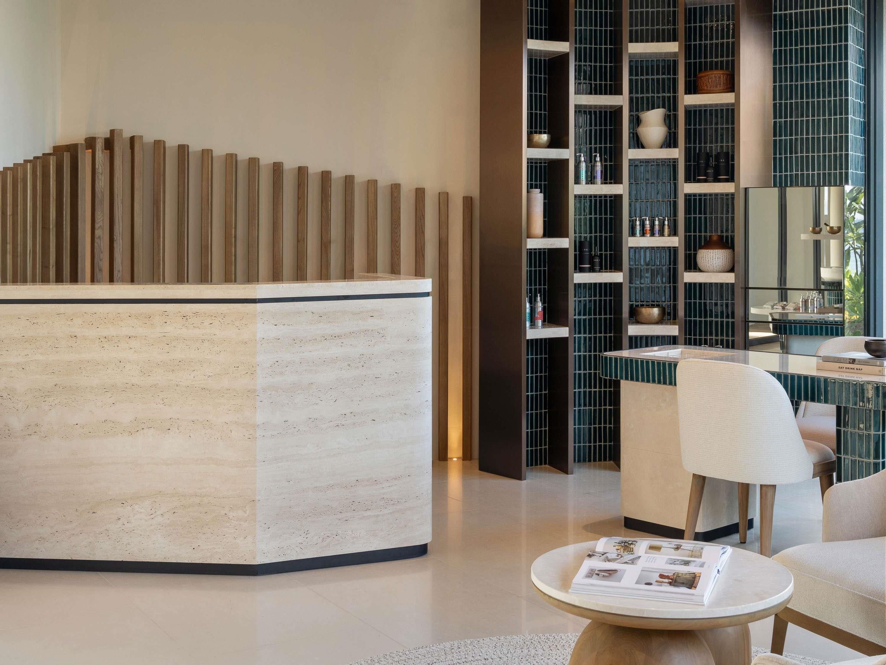 Uma área de recepção com um balcão de mármore, um espaço de espera com uma mesa redonda e uma estante com diversos objetos.