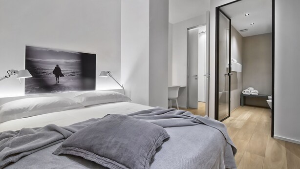Quarto moderno com uma cama grande, uma mesa de cabeceira, uma luminária de parede e um banheiro visível através de uma porta de vidro.
