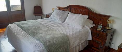 Cama bem arrumada com colcha acolchoada e travesseiros, mesa de cabeceira de madeira com abajur, e uma cadeira num quarto com chão ladrilhado e porta com painel de vidro.