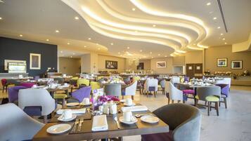 Ampla sala de jantar de hotel, com várias mesas e cadeiras, design moderno no teto e serviço de buffet.