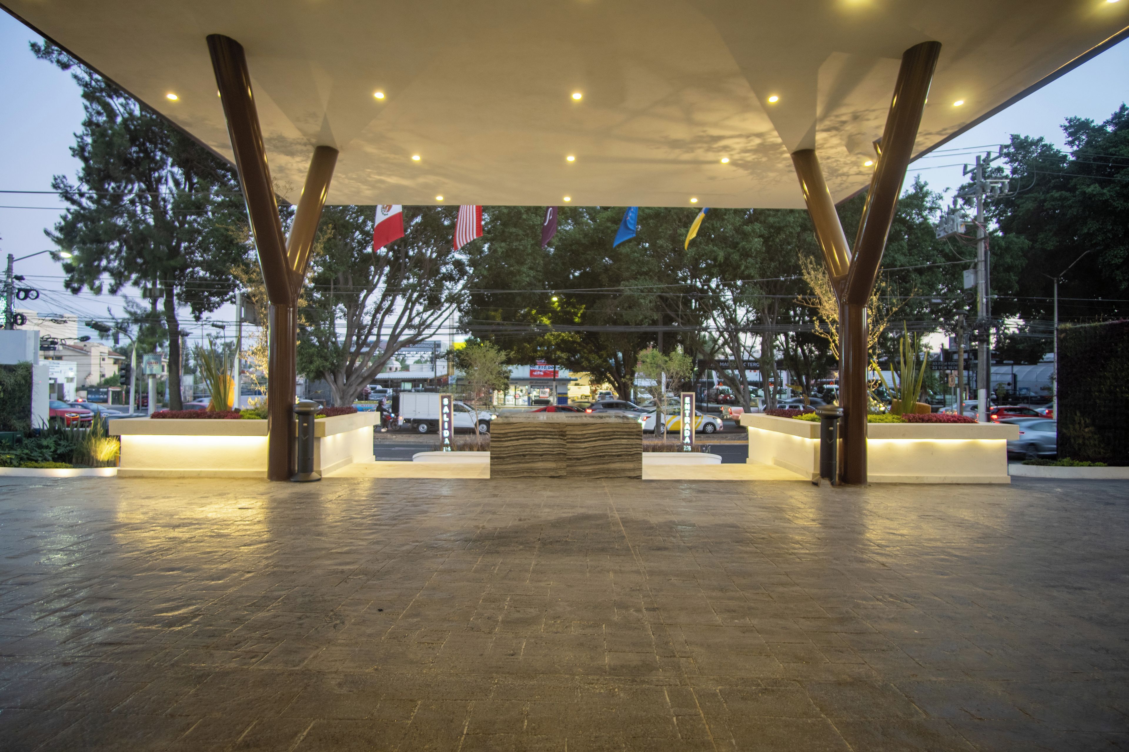 Área externa coberta com um espaço central para assentos, rodeada por árvores e um estacionamento ao fundo.