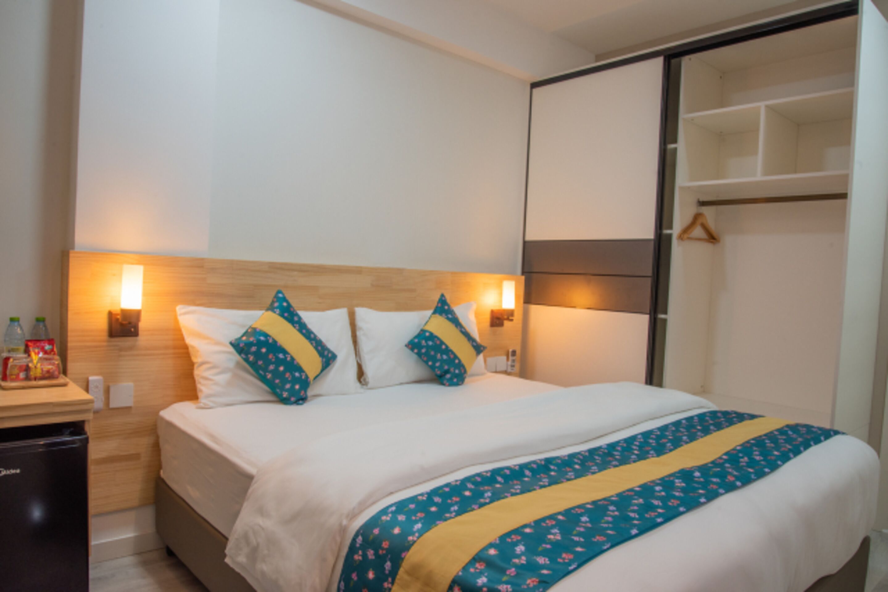 Cama bem arrumada com manta de padrão floral, cabeceira de madeira e luminárias laterais.