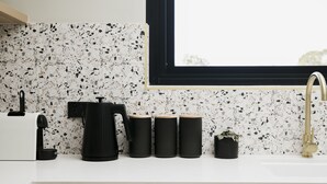 Cozinha com bancada branca, máquina de café preta, três recipientes pretos, uma pequena planta em vaso e uma torneira dourada.
