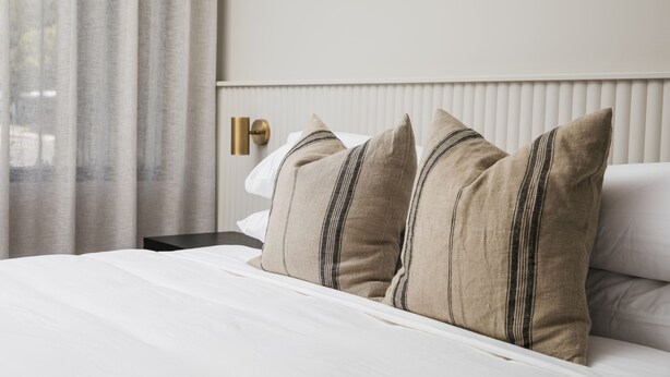 Uma cama bem arrumada com lençóis brancos e almofadas bege com listras pretas, uma mesa de cabeceira com um objeto dourado e uma janela com cortinas finas.
