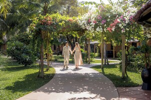 Um casal caminhando sob um arco de flores em um jardim, com um prédio de tijolos ao fundo.