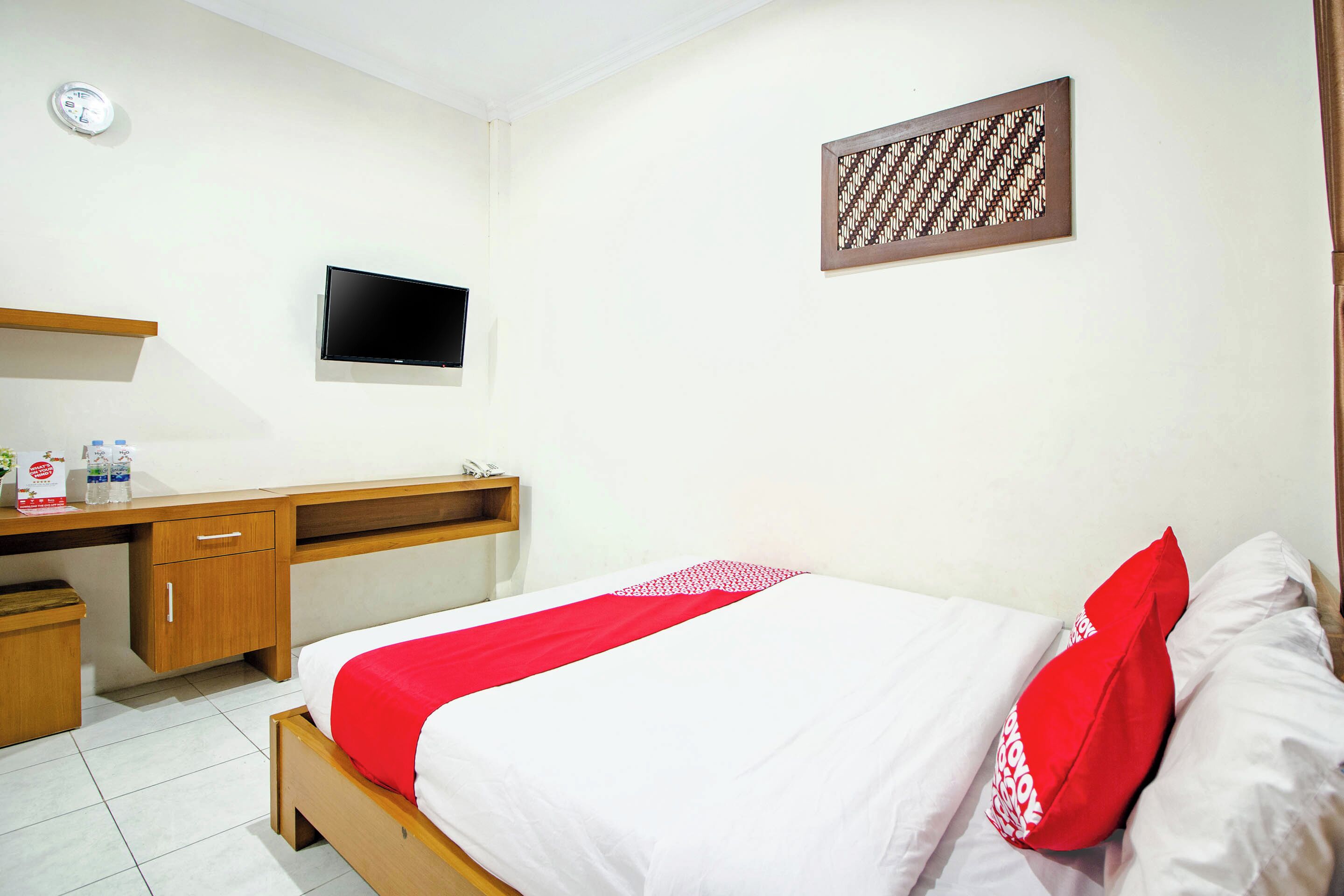 Quarto de hotel com uma cama, uma escrivaninha com cadeira, uma televisão e uma pequena prateleira com objetos.
