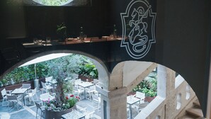 Uma área de refeições ao ar livre com mesas e cadeiras, uma grande planta em vaso e vista do jardim através de um arco.