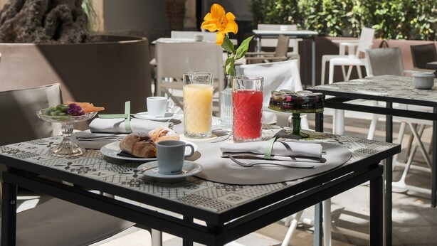 Uma área de refeições ao ar livre com mesas preparadas para o café da manhã, incluindo croissants e frutas.