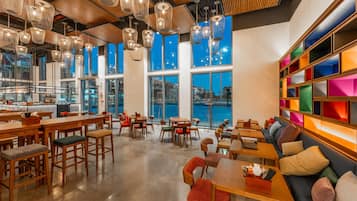 Um café espaçoso com pé-direito alto, luminárias de vime suspensas e vista para a paisagem urbana através de grandes janelas.