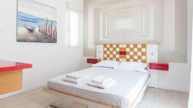 Quarto moderno com cabeceira estampada em xadrez, cama com toalhas brancas bem dobradas e mesa de cabeceira vermelha.
