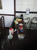 Uma mesa de jantar com diversos itens, incluindo uma caixa de TIROL, uma cafeteira, uma cesta de pão e várias chávenas e tigelas.