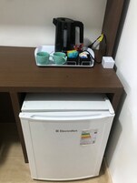 Uma geladeira branca compacta com painel de controle e um adesivo, uma escrivaninha marrom com uma cafeteira preta, uma bandeja com duas xícaras verde-água e uma caixa pequena, e um régua de energia branca.