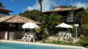 Uma área de piscina com cadeiras de lounge brancas e guarda-sóis, rodeada por vegetação exuberante e um edifício com telhado em azulejos.