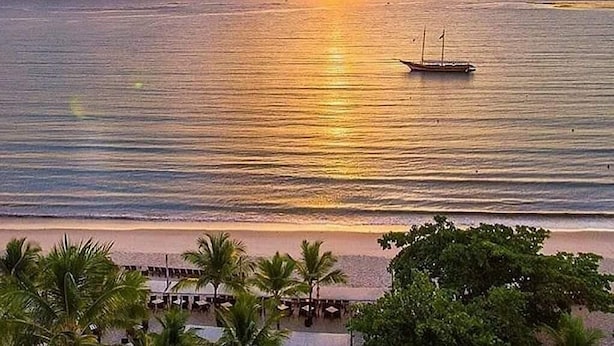 Um pôr do sol sobre um mar tranquilo, com um barco e palmeiras em primeiro plano.