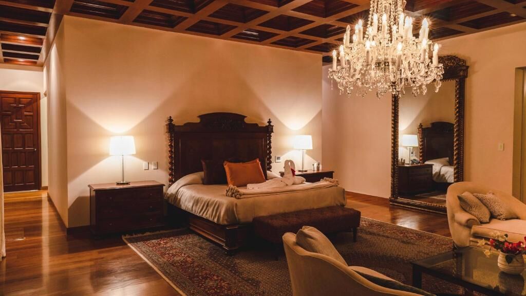 Quarto com cama grande, duas mesas de cabeceira, um lustre, um espelho e um espaço de estar com uma cadeira e mesa.