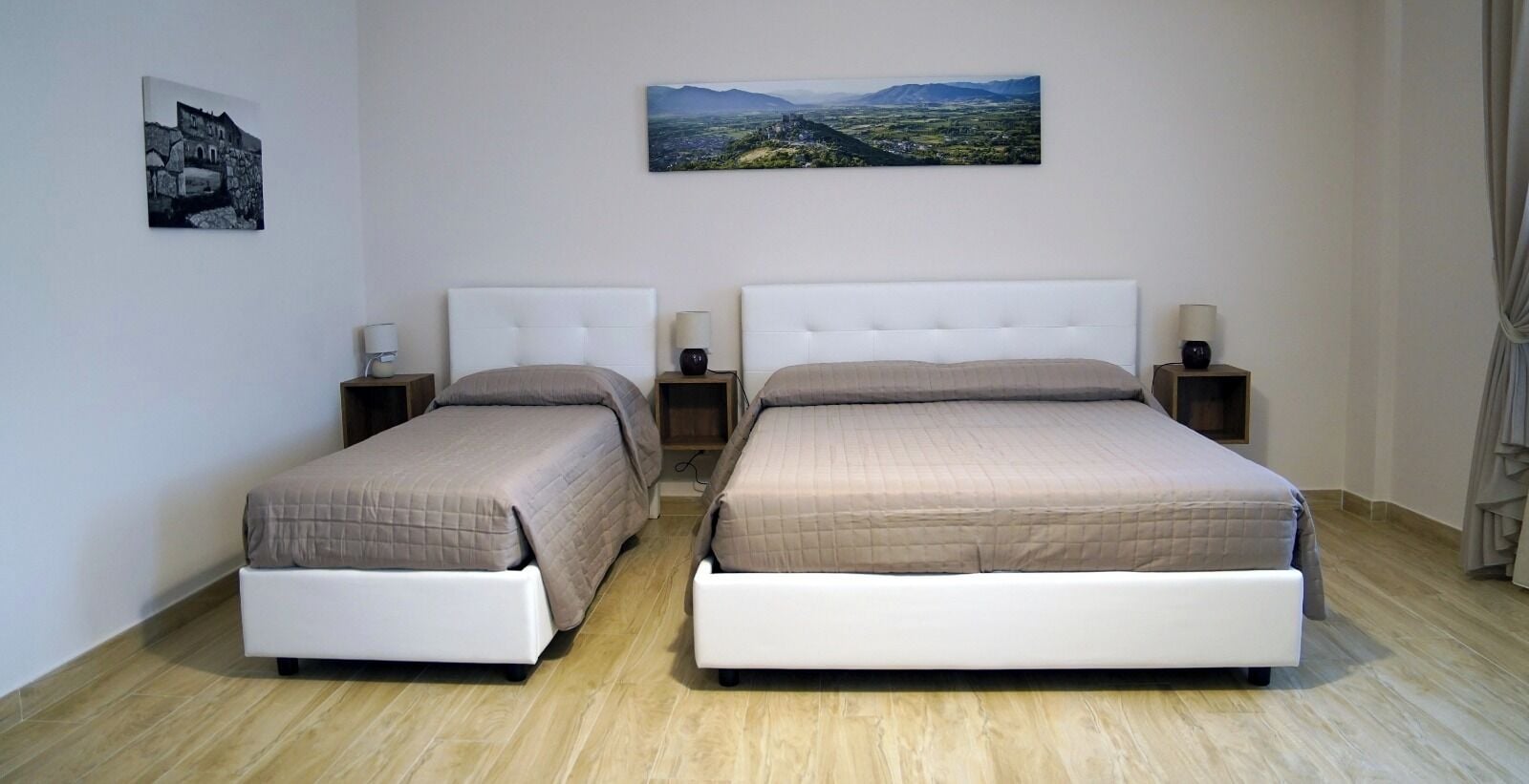 Quarto duplo com duas camas, cada uma com cabeceira branca e colcha bege acolchoada. Há duas mesas de cabeceira com abajures, e um quadro de paisagem pendurado acima das camas.