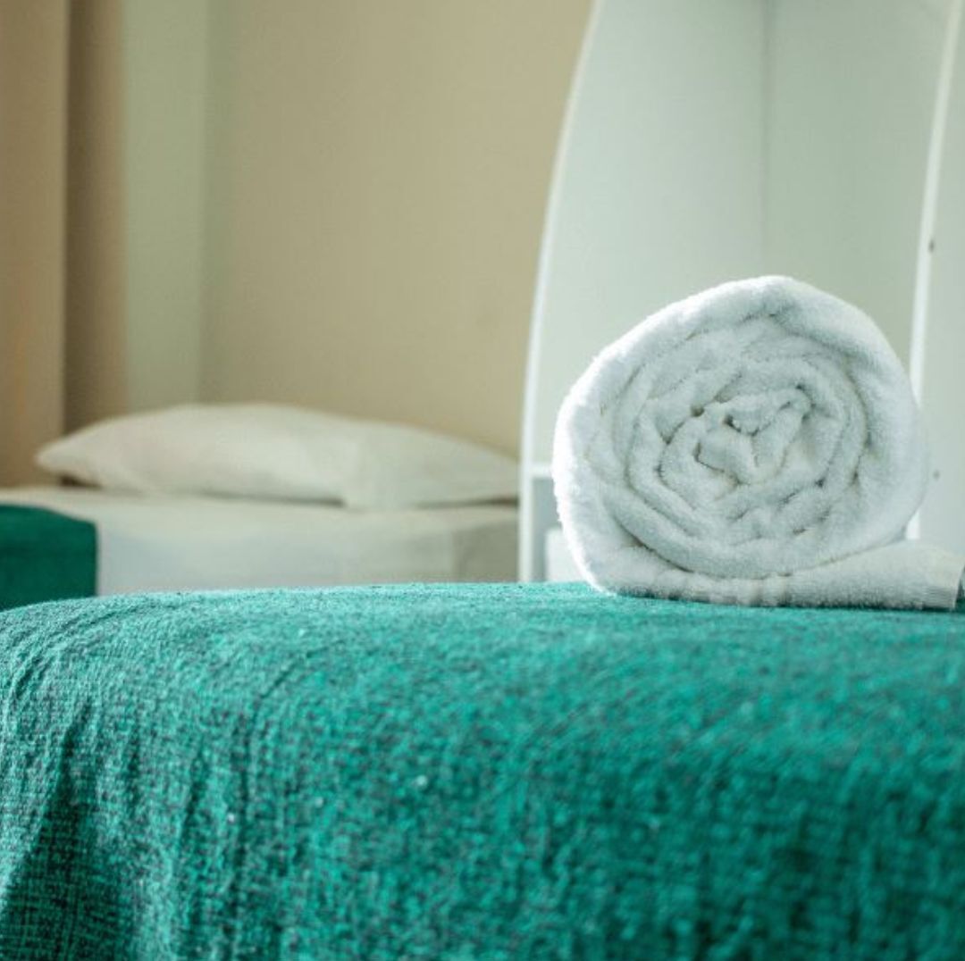 Cama bem arrumada com manta azul-turquesa e toalha branca enrolada sobre um travesseiro branco.