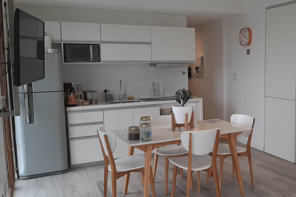 Uma cozinha compacta com armários brancos, micro-ondas, torradeira e uma mesa de jantar com quatro cadeiras.