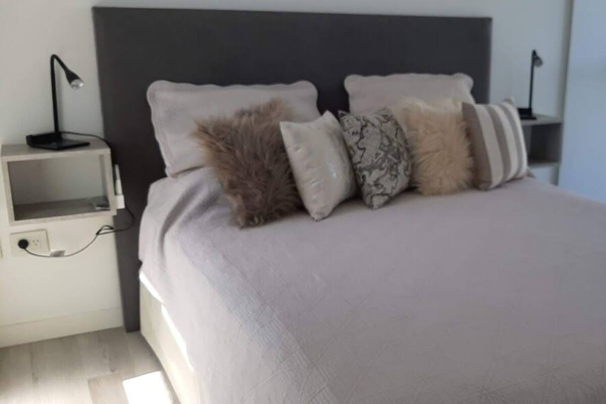 Cama bem arrumada com cabeceira cinza, mesa de cabeceira com abajur e janela com vista para edifícios e árvores.