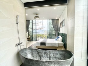 Uma casa de banho moderna com uma banheira grande de apoio, uma área de duche com paredes de vidro e uma cama com vista para o exterior.