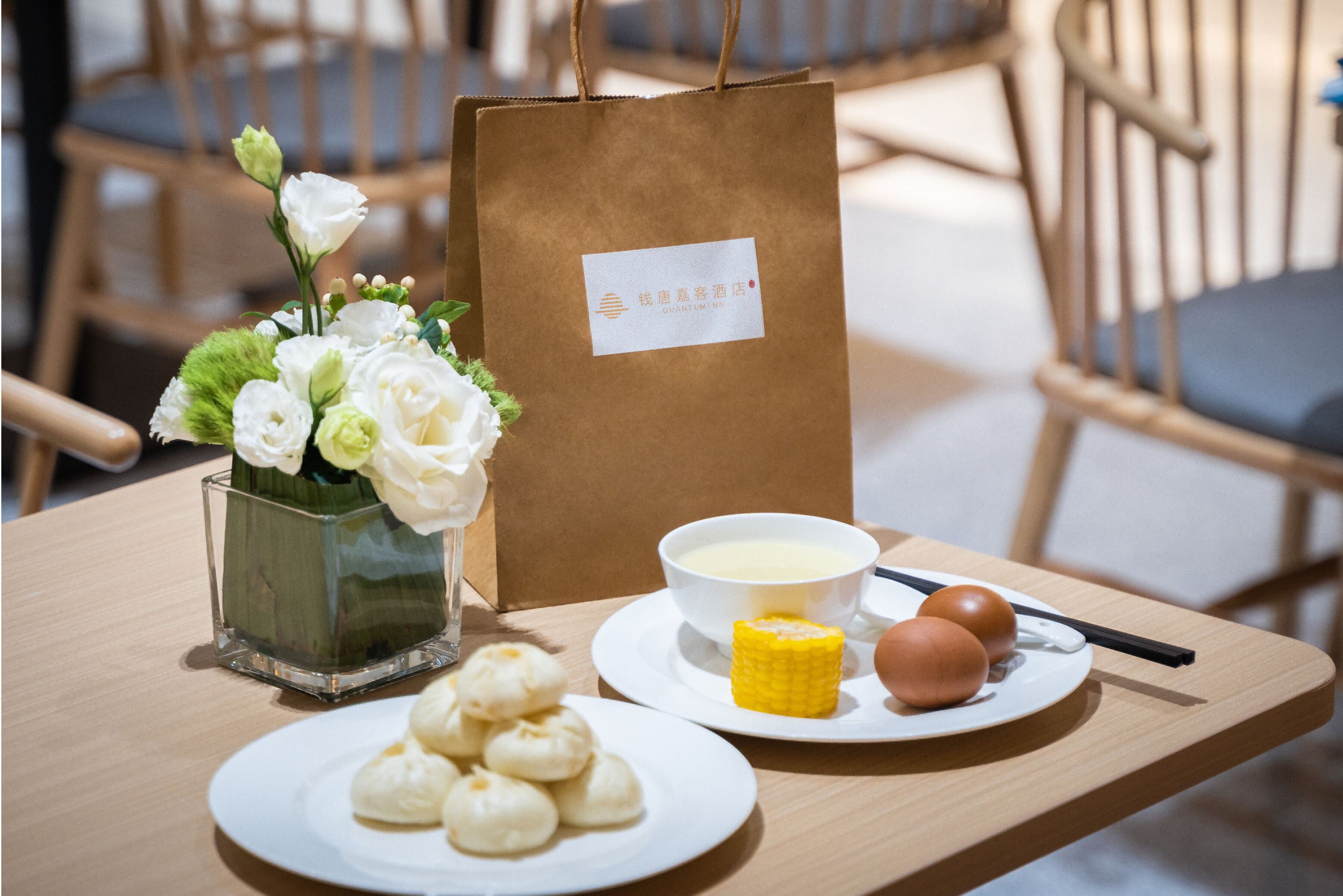 Uma mesa com um saco de papel castanho, uma tigela de sopa, um prato com milho e ovos, e um vaso com flores brancas.