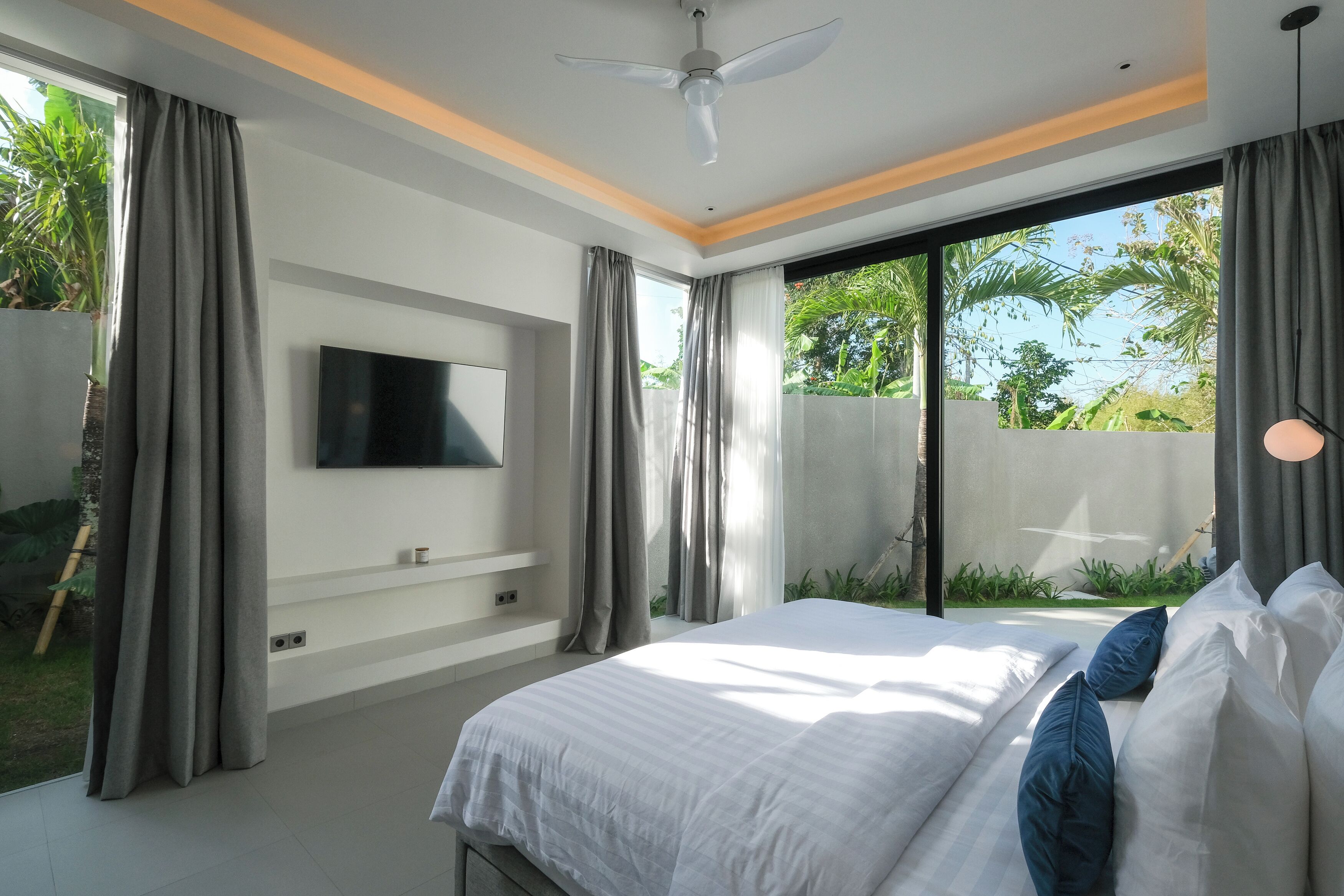 Quarto moderno com cama grande, ventoinha no teto, TV fixada na parede e vista para o verde lá fora.