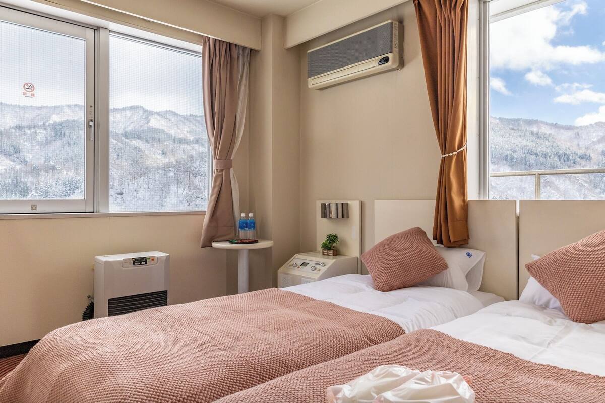 Quarto de hotel com duas camas, mesa de cabeceira com luminária, janela com cortinas, ar condicionado e vista para montanhas cobertas de neve.
