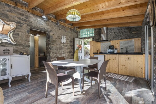 Interior rústico com parede de pedra, teto de madeira e uma mesa de jantar redonda com cadeiras.