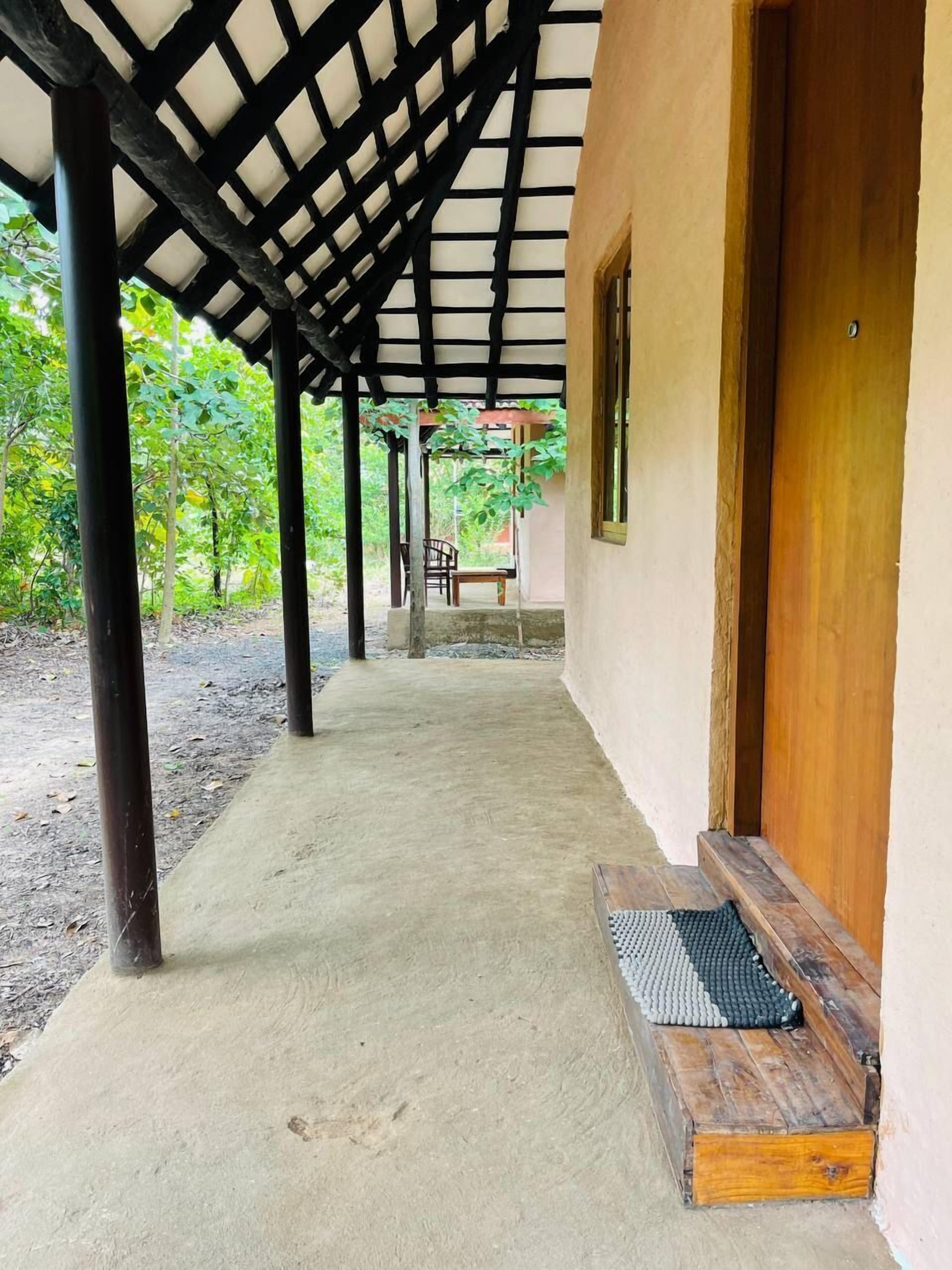 Uma área externa coberta com piso de concreto, degraus de madeira que levam a uma porta e um banco sob uma pérgola.