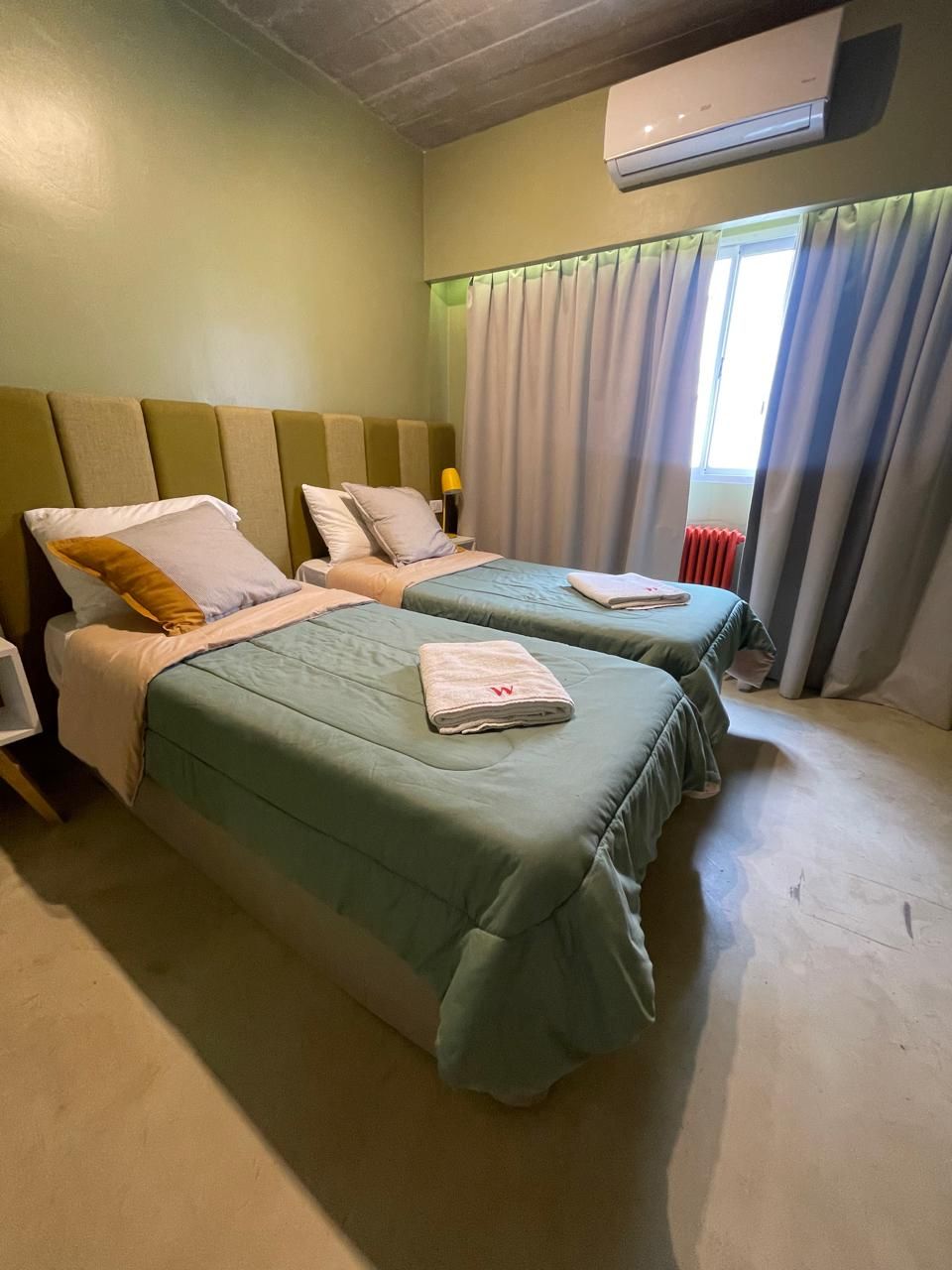 Quarto de hotel com duas camas, uma janela com cortinas, um aparelho de ar condicionado e uma luminária na mesa de cabeceira.