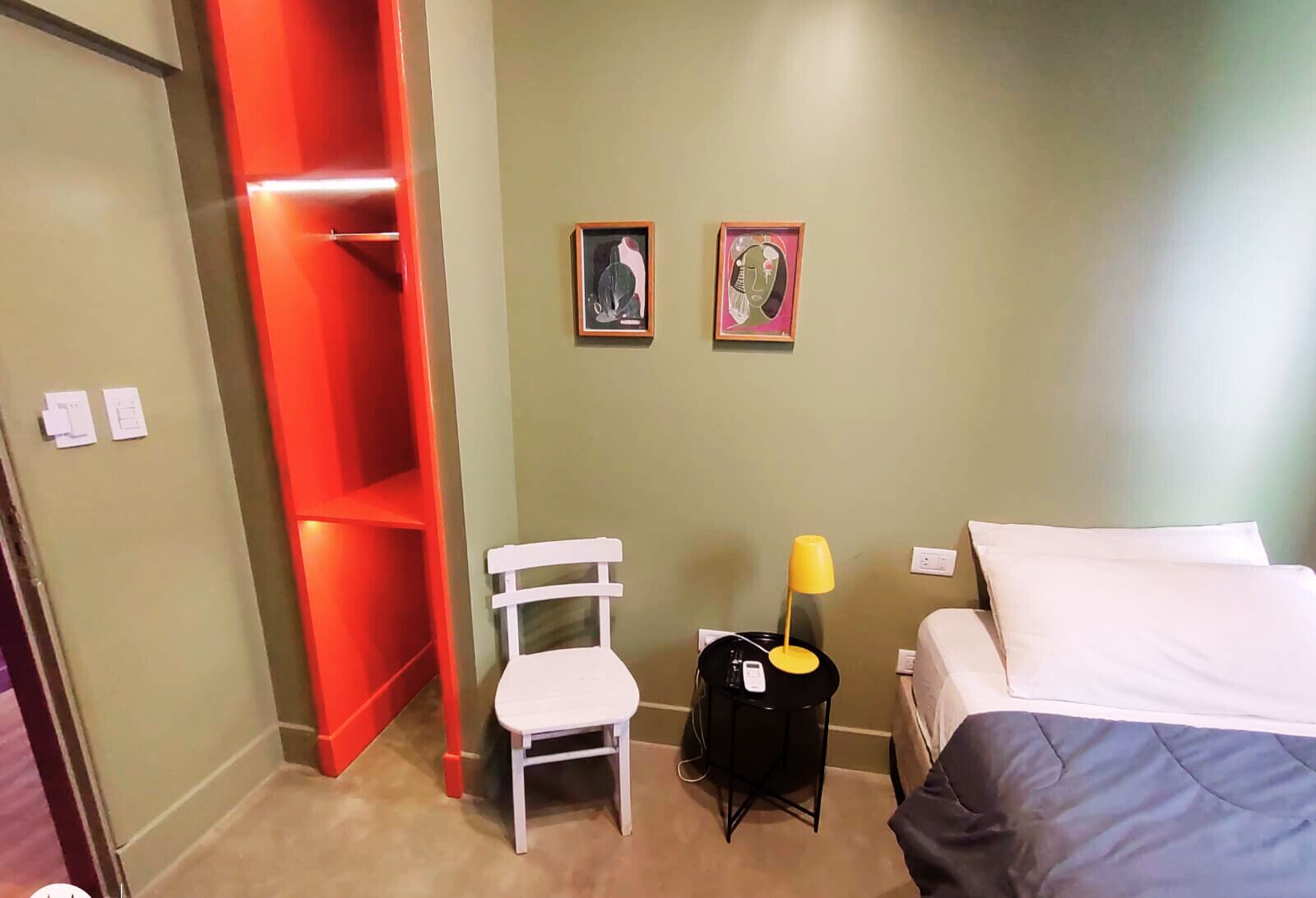 Quarto com uma cama, uma cadeira, uma mesinha de cabeceira com abajur, e duas quadros na parede.