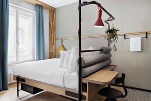 Quarto de hotel moderno com cama, escrivaninha, cadeira e planta pendente.