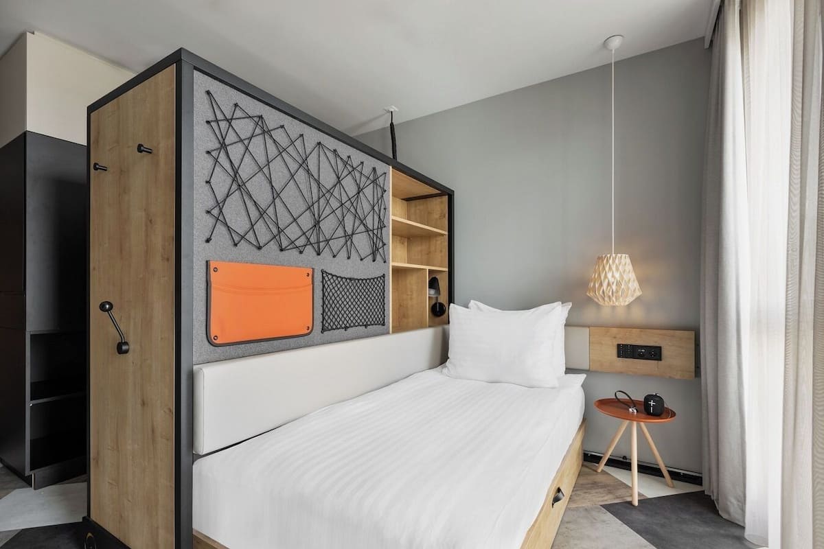 Quarto de hotel moderno com uma cama grande, um guarda-roupa de madeira, uma escrivaninha com abajur e uma mesa de cabeceira.