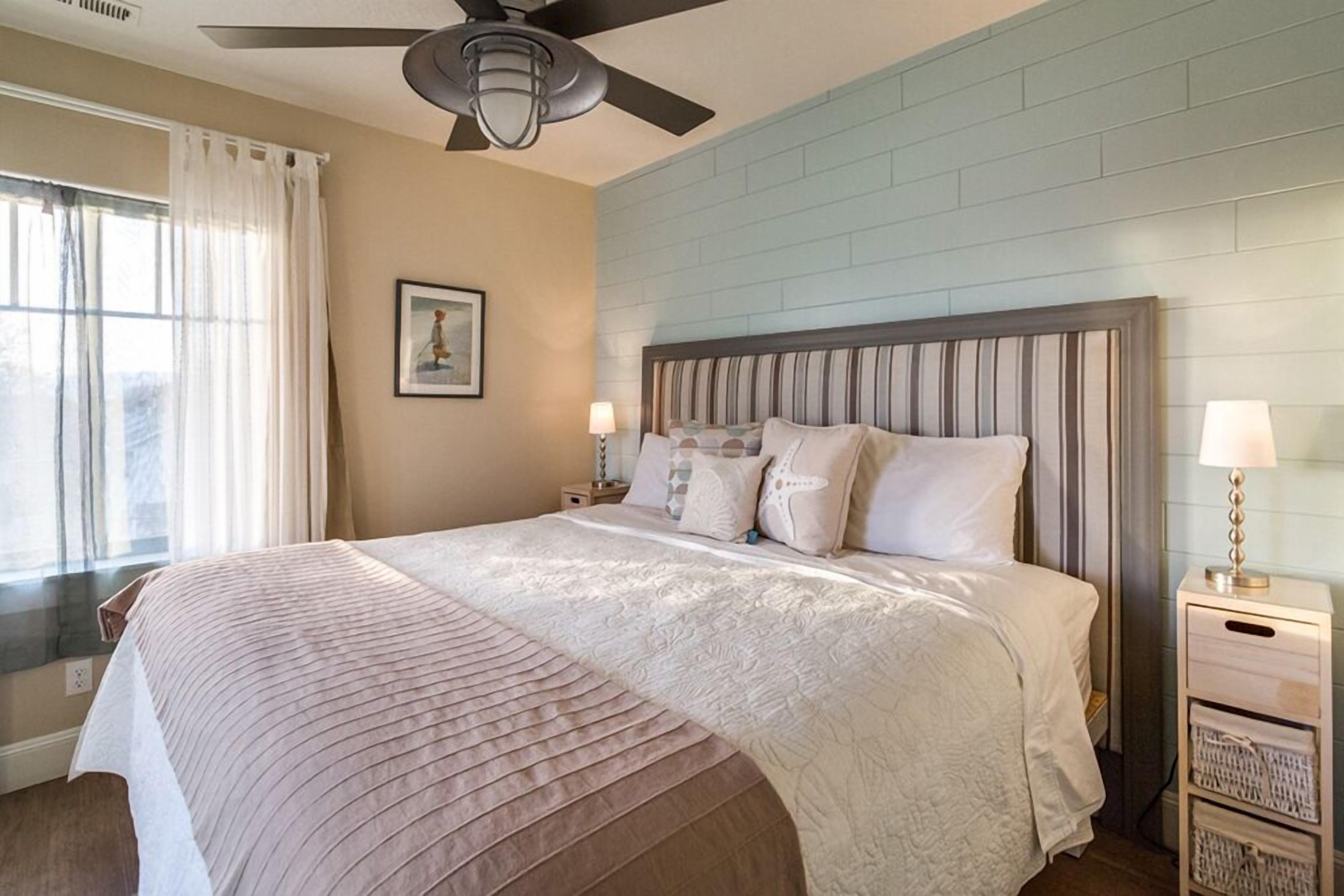 Quarto com cama grande, ventoinha no teto, janela com cortinas e mesa de cabeceira com abajur.