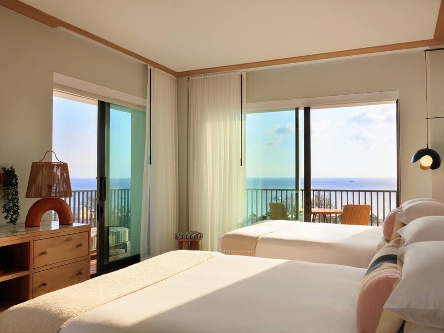 Quarto de hotel com duas camas, cómoda de madeira, televisão e varanda vista para o oceano.