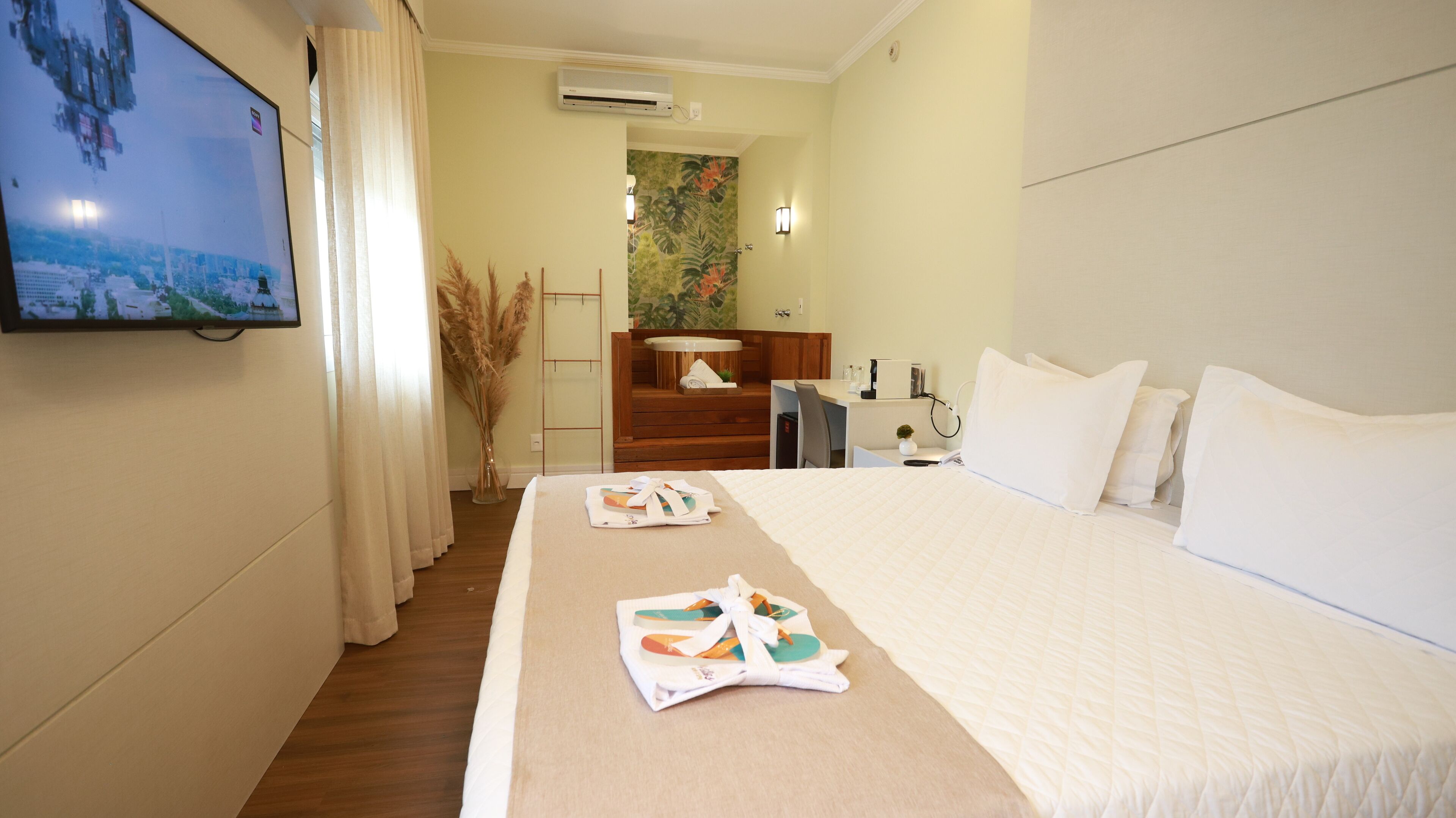 Quarto de hotel com cama grande, escrivaninha, cadeira, televisão e banheiro com chuveiro e vaso sanitário.