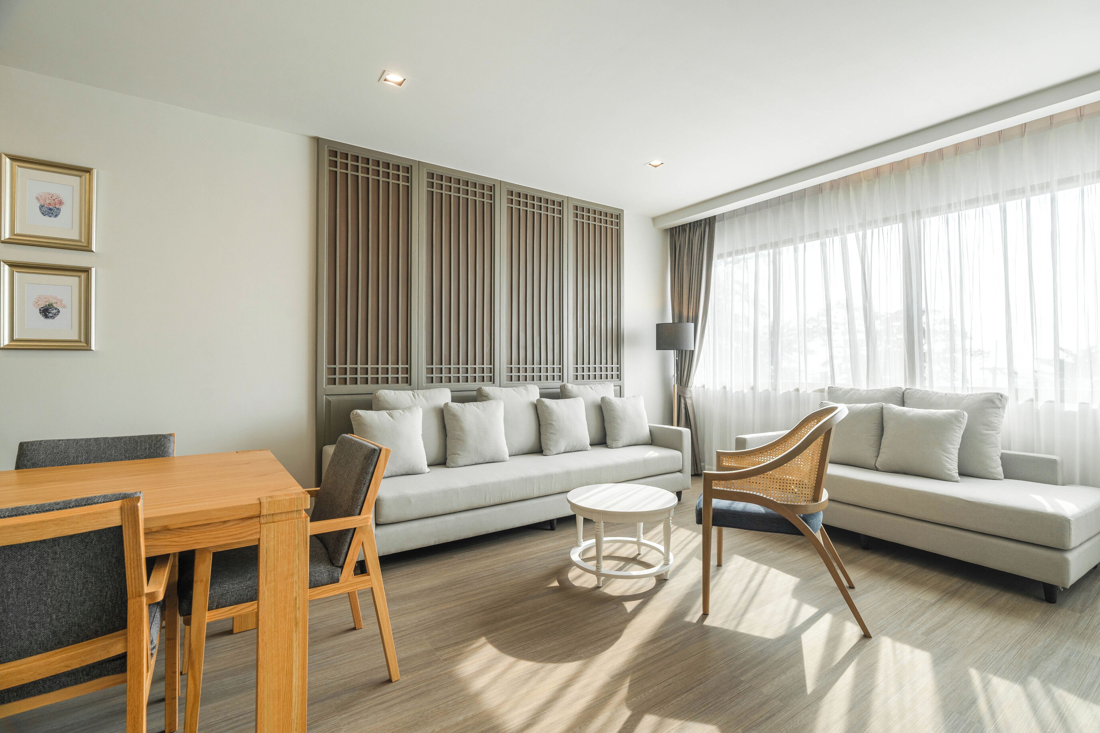 Uma sala de estar moderna com um sofá, uma mesa de jantar com cadeiras, uma mesa de centro redonda e uma janela grande com cortinas leves.