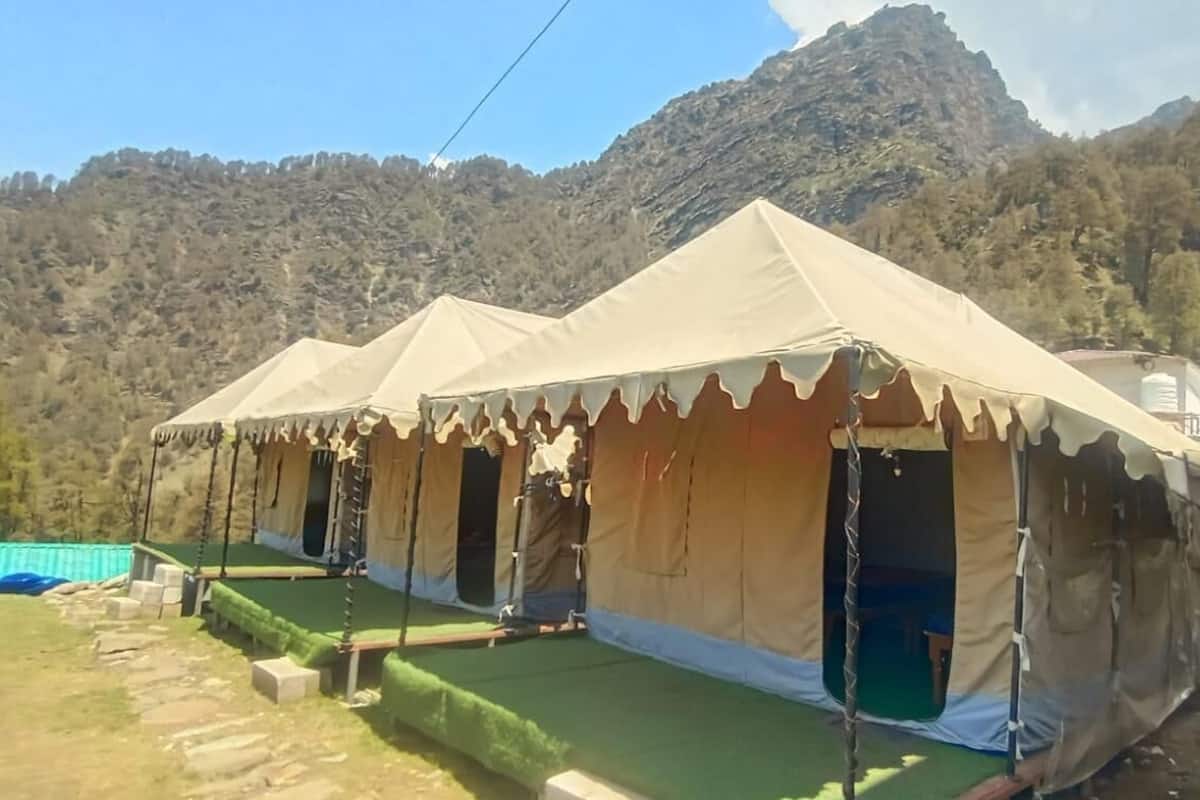 Fileira de tendas bege com carpetes verdes e uma parede de pedra, ao fundo montanhas.