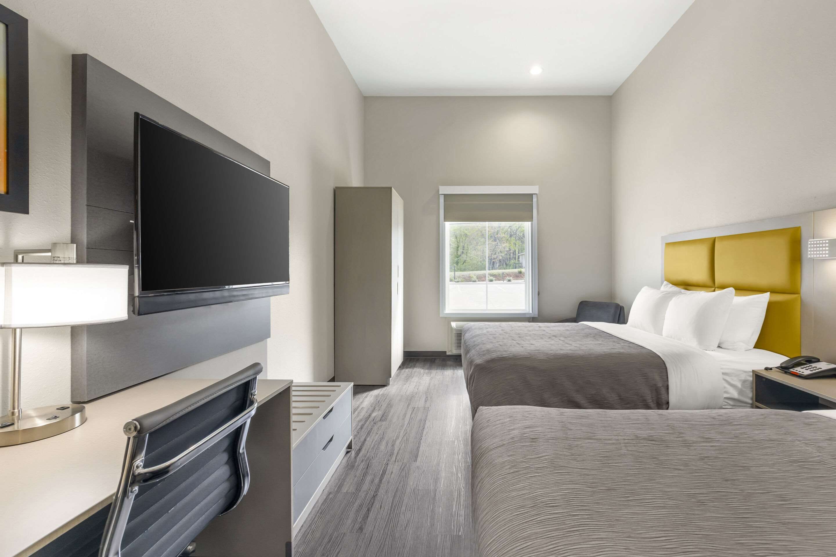 Quarto de hotel moderno com televisão de tela plana, duas camas, mesa de cabeceira, abajur e janela com vista.
