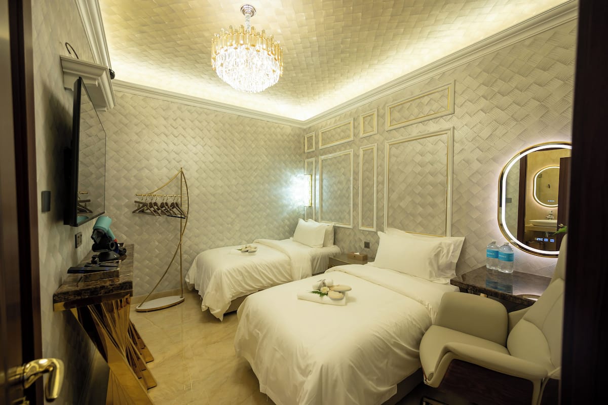 Quarto de hotel com duas camas, um candelabro, um espelho e uma pequena mesa com uma garrafa de água.