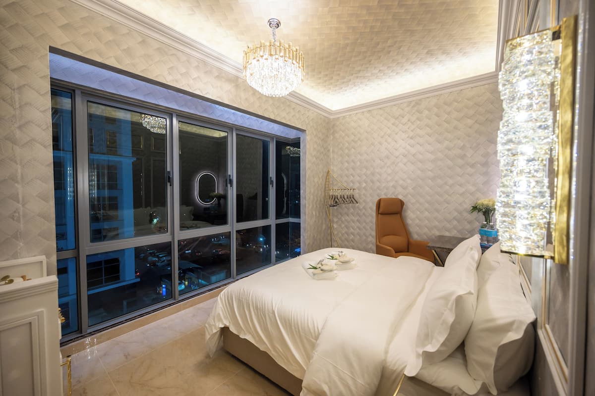 Quarto com uma janela grande, uma cama, um candelabro e vista da cidade à noite.