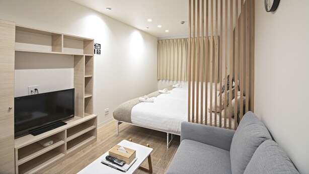 Quarto de hotel moderno com televisão de tela plana, sofá, cama com cabeceira de madeira e divisória de madeira deslizante.