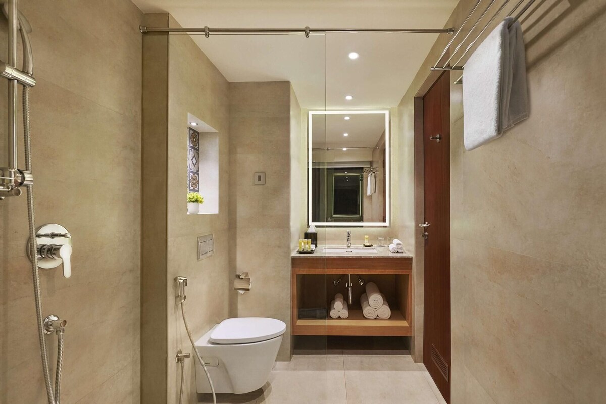 Uma casa de banho moderna com chuveiro em cabine de vidro, sanita branca, toucador de madeira com espelho e um porta-toalhas.