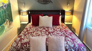 Cama bem arrumada com colcha de padrão vermelho e branco, duas almofadas listradas e uma almofada xadrez vermelha. Duas luminárias de cabeceira em cada lado da cama, e uma pintura de paisagem montanhosa visível na parede à esquerda.