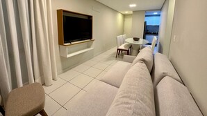 Uma sala de estar moderna com televisão de tela plana na parede, uma mesa de jantar redonda com cadeiras e um sofá bege grande.