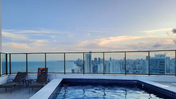 Piscina no terraço com vista para a cidade, cadeiras de descanso e piso ladrilhado.